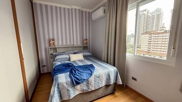 Quarto com cama, cortinas listradas, janela com vista para a cidade e um aparelho de ar condicionado.