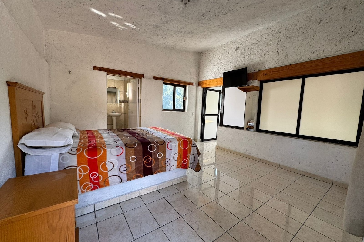 Quarto com cama, cabeceira de madeira, escrivaninha, banheiro visível por uma porta aberta e uma janela grande.
