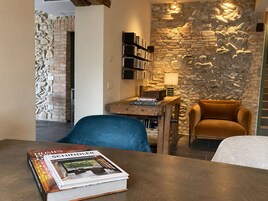 Uma sala de estar acolhedora com uma parede de pedra, estante de livros, uma poltrona azul e uma mesa com uma revista.