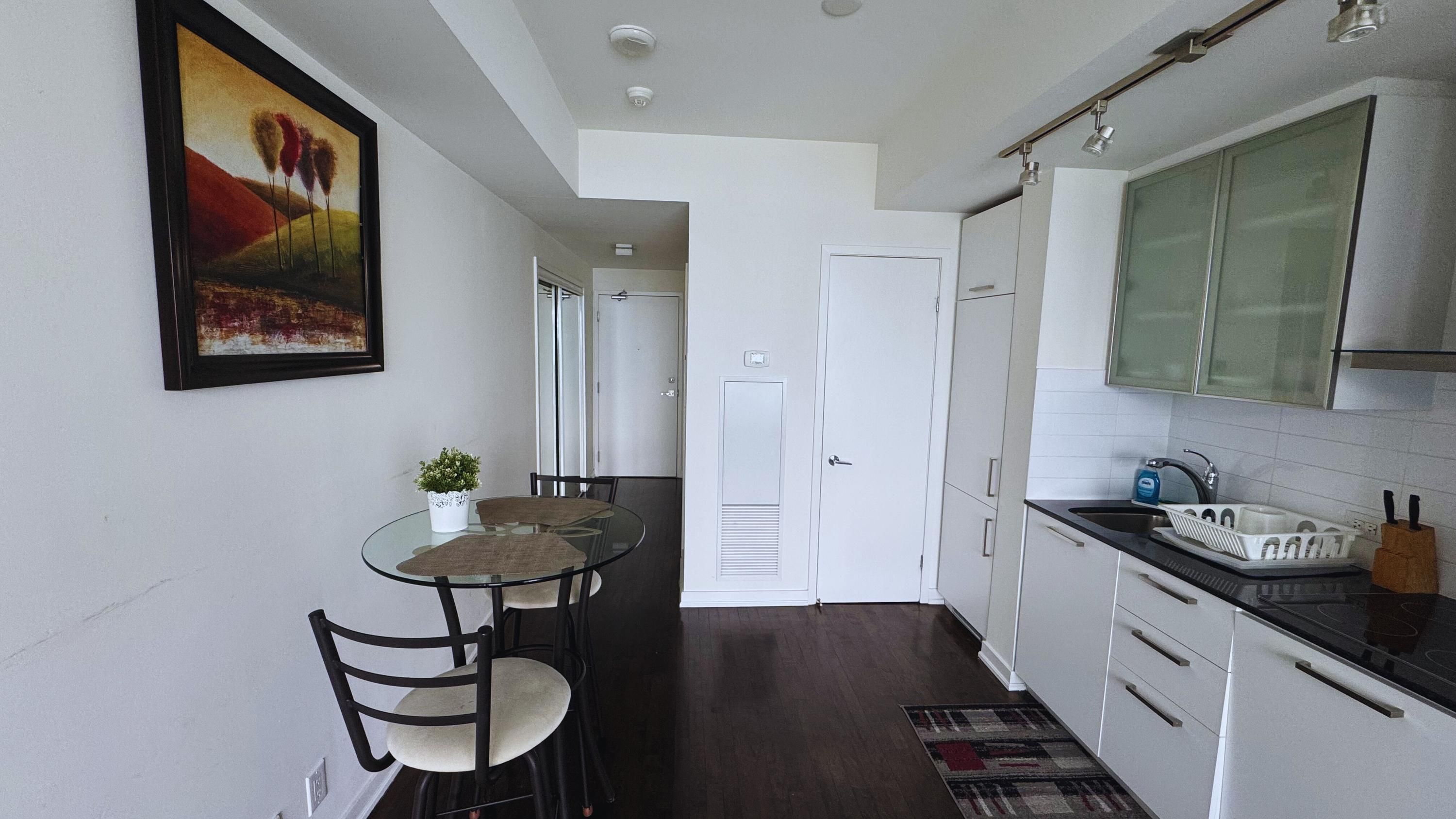 Uma cozinha compacta com armários brancos, bancada preta e um espaço de refeições com mesa e cadeiras de vidro.