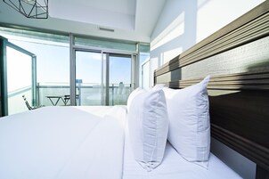 Quarto de hotel com uma cama grande, roupa de cama branca, cabeceira de madeira e uma varanda com mesa e cadeiras.