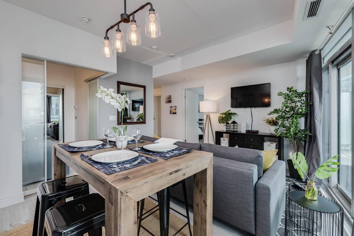 Uma sala de estar moderna com uma mesa de jantar de madeira posta para dois, um sofá cinzento e uma grande janela com vista.