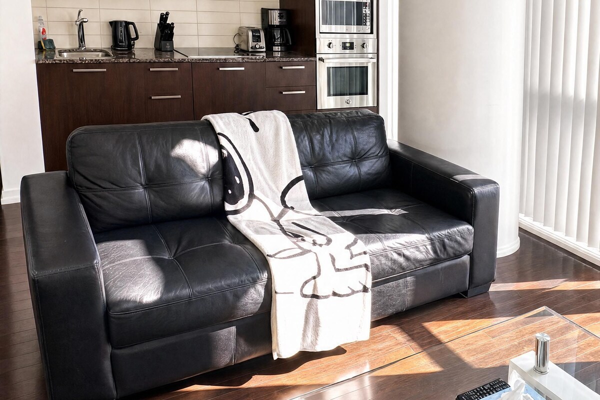 Uma sala de estar moderna com um sofá de couro, uma mesa de centro de vidro e uma cozinha ao fundo.