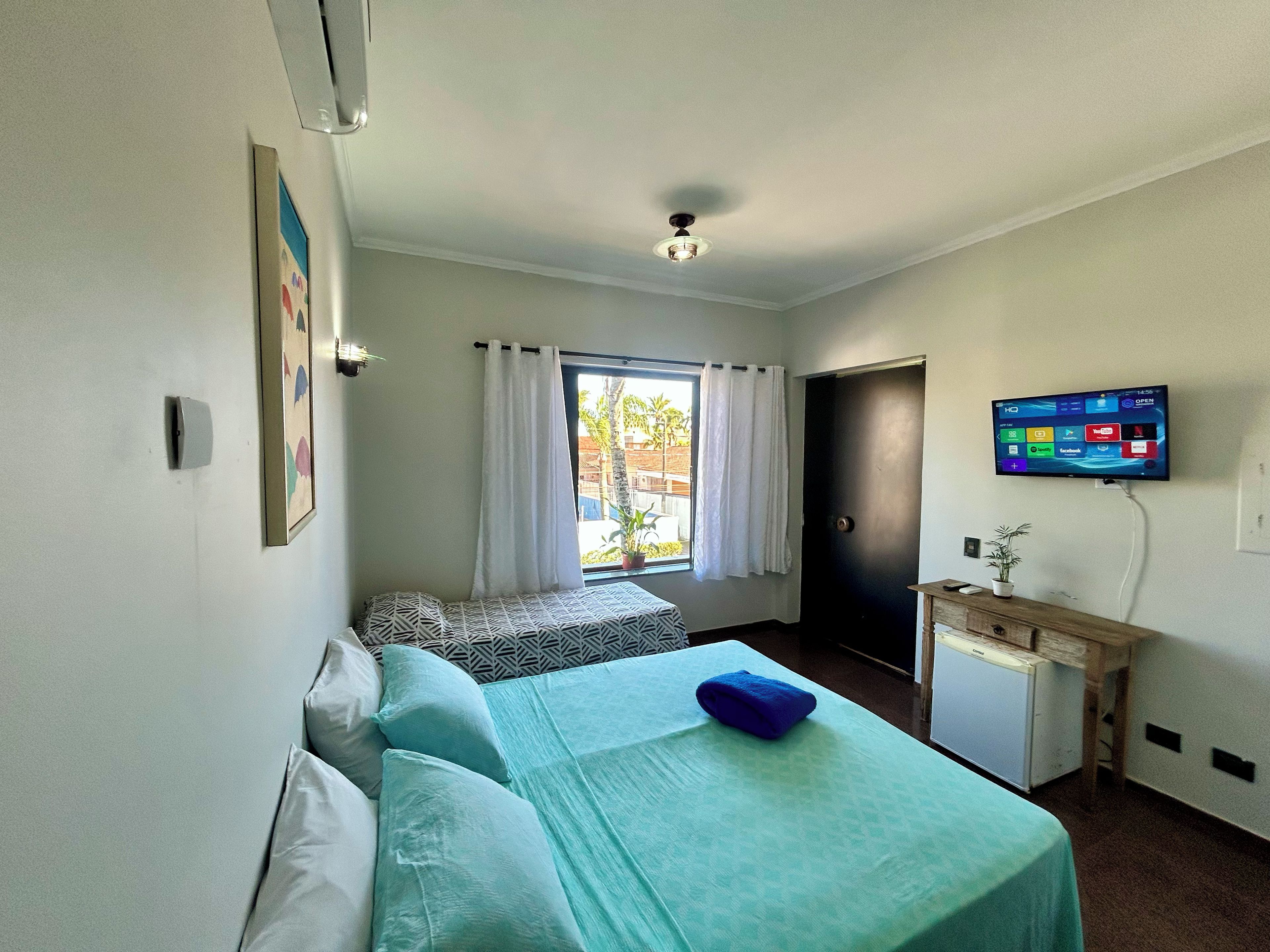 Quarto com duas camas, uma escrivaninha, televisão e janela com cortinas.