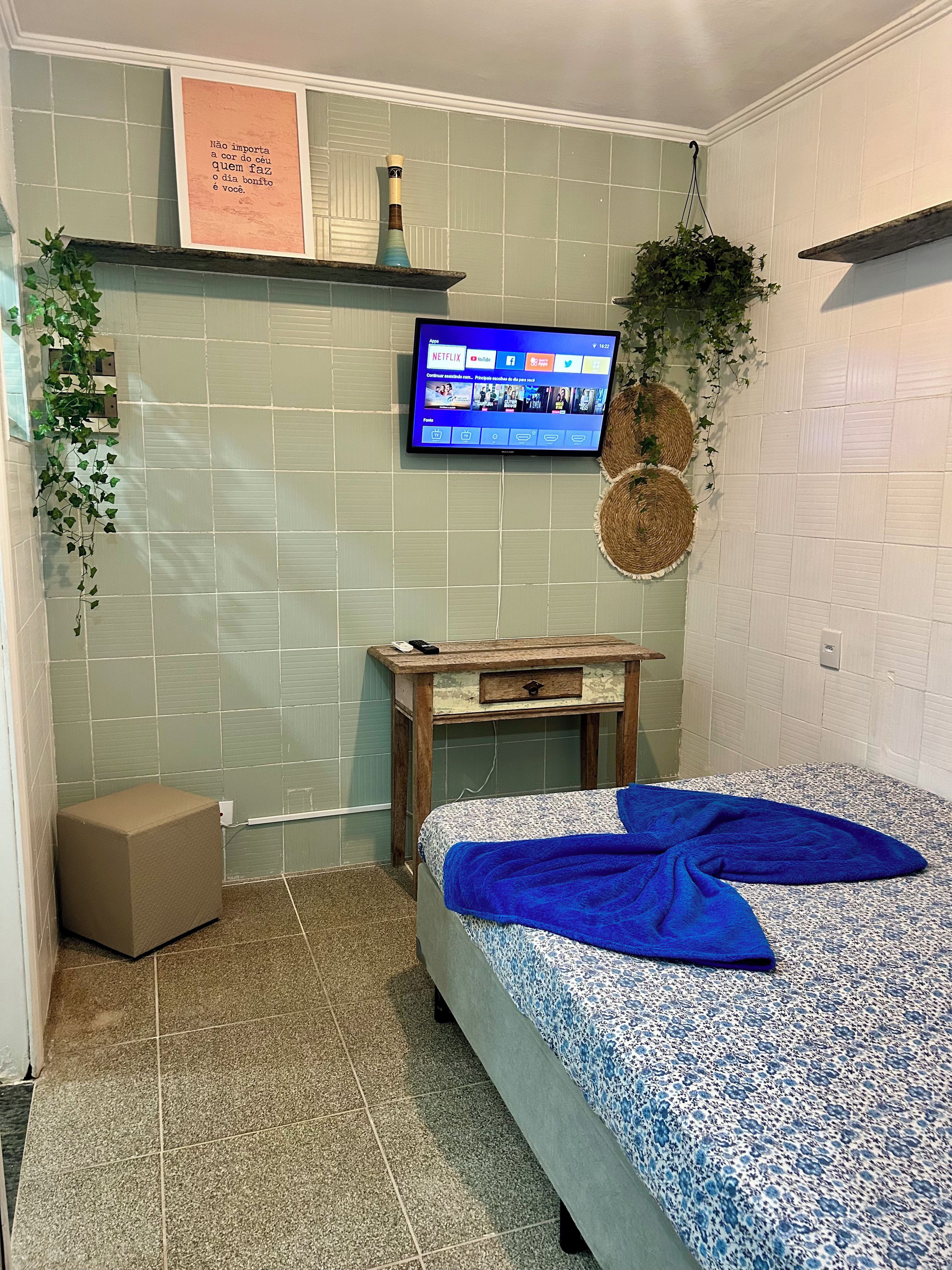 Quarto com cama, televisão na parede, pequena mesa de madeira e prateleira com planta.