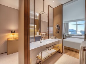 Quarto de hotel moderno com uma cama grande, banheiro com toucador, espelho e vista da cidade.