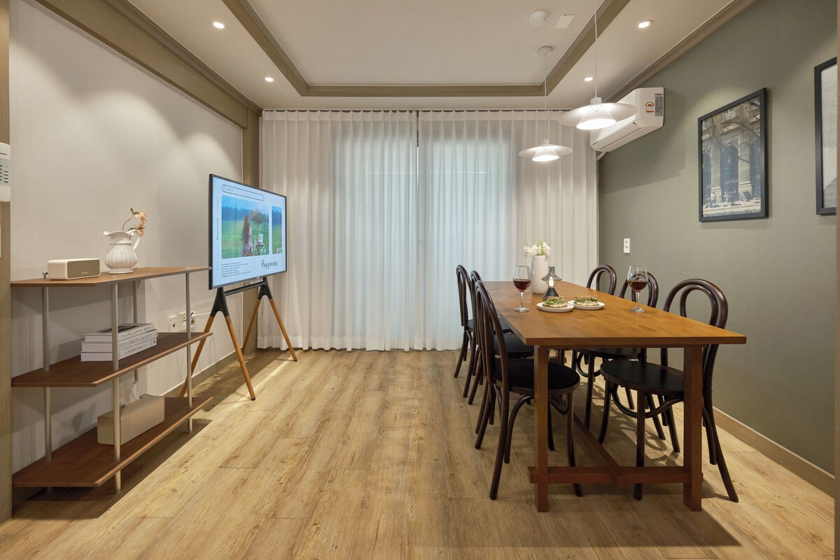 Sala de conferências com mesa de madeira, cadeiras, tela de projetor e cortina branca.