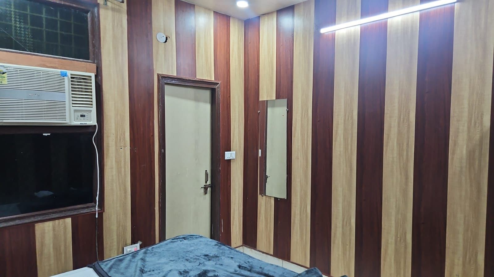 Quarto com paredes de madeira listrada, uma porta, um espelho pequeno e uma cama com cobertor azul.