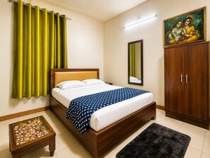 Quarto de hotel com cama, armário de madeira, pequena mesa com toalha de mesa florida e um quadro na parede.