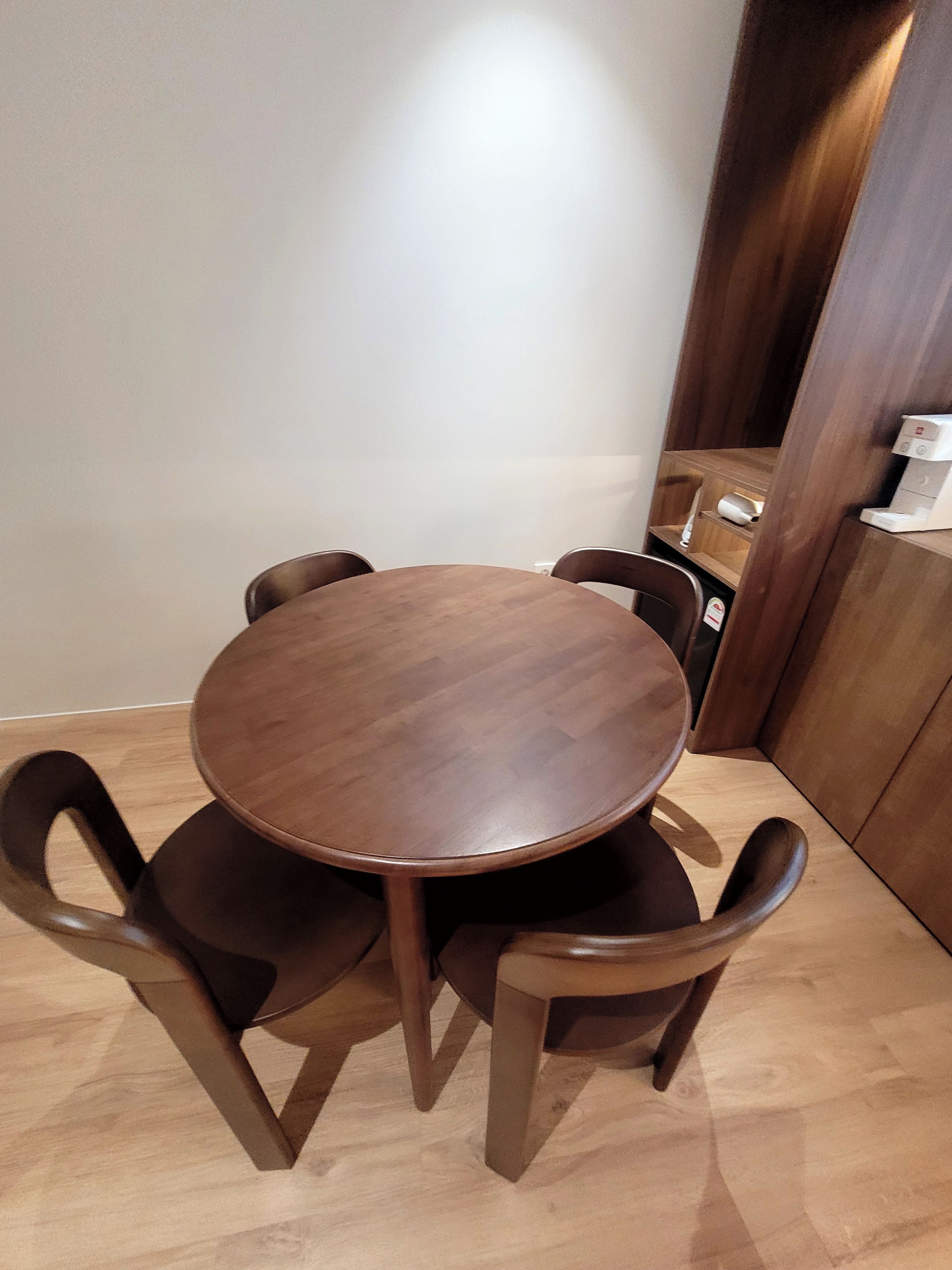 Mesa redonda de madeira com quatro cadeiras, uma máquina de café e um armário de madeira em um quarto com piso claro.