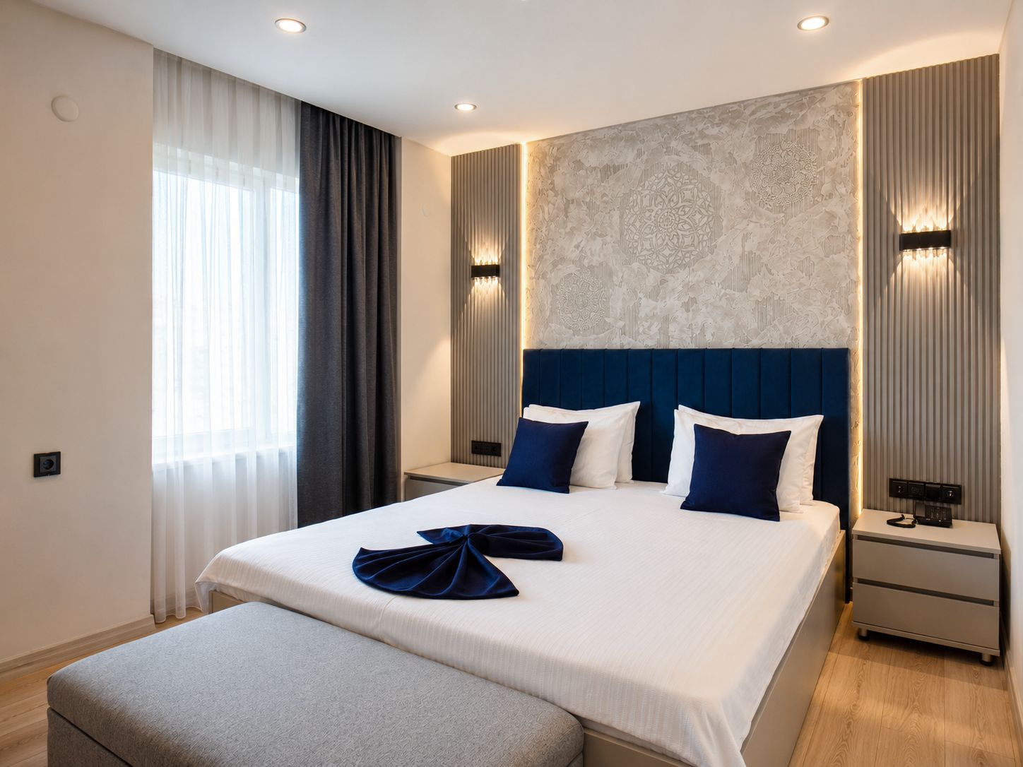 Quarto de hotel com cama grande, mesa de cabeceira, rodapé e janela com cortinas.
