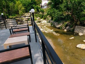Um deck à beira do rio com mesas e cadeiras de madeira, corrimão e vista para um rio com pedras e vegetação.