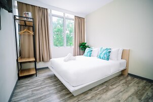 Cama bem arrumada com lençóis brancos e duas almofadas decorativas, uma prateleira de madeira com uma peça de roupa pendurada e uma janela com cortinas.