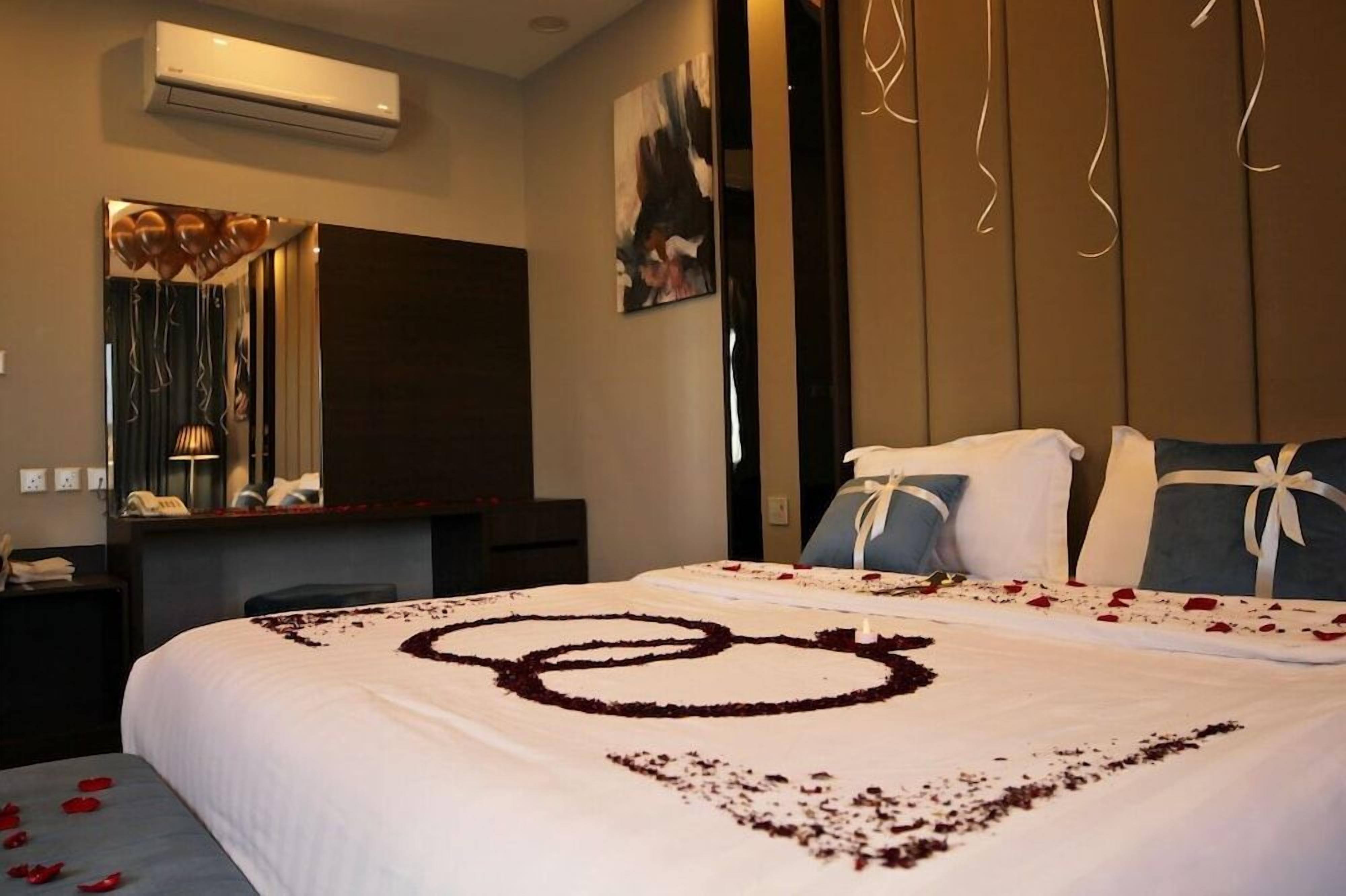 Quarto de hotel com uma cama decorada com pétalas de rosa e um desenho em formato de coração, uma mesa com abajur e um ar-condicionado fixado na parede.