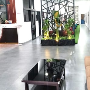 Uma área de recepção com balcão, uma mesa de vidro com planta e um espaço de estar verde.