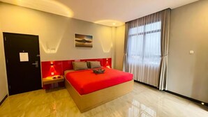Quarto de hotel com cama grande, mesa de cabeceira, cabeceira vermelha, quadro na parede e janela com cortinas.