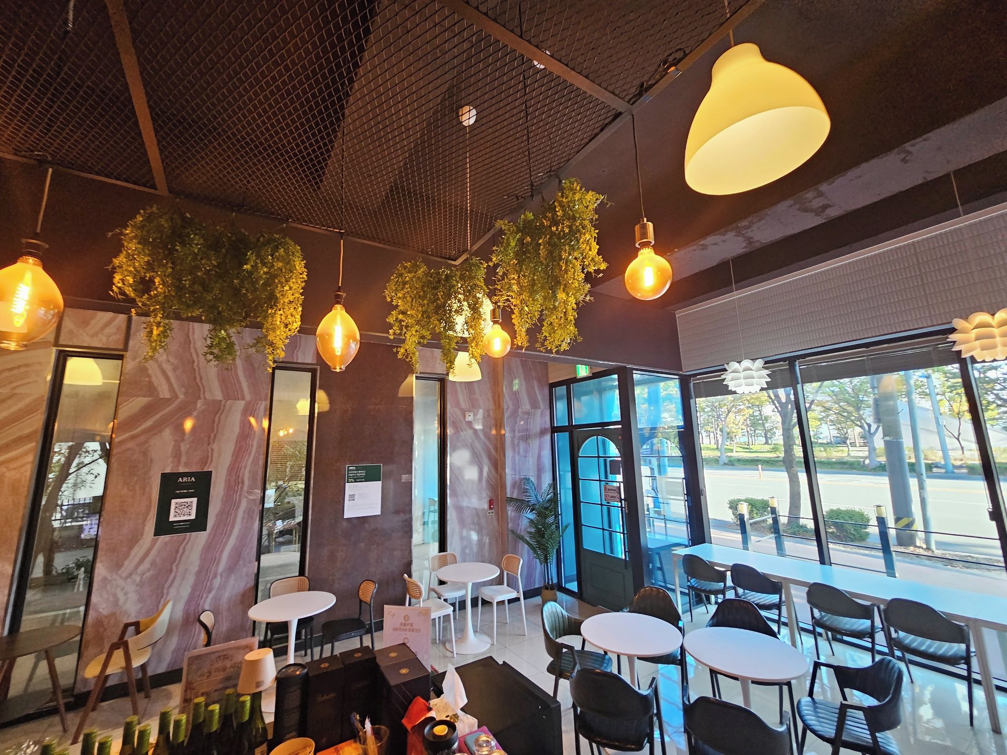 Interior de restaurante com plantas penduradas, luminárias pendentes e vista para a rua através de grandes janelas.