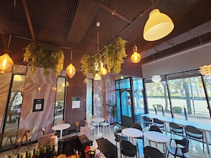 Interior de restaurante com plantas penduradas, luminárias pendentes e vista para a rua através de grandes janelas.