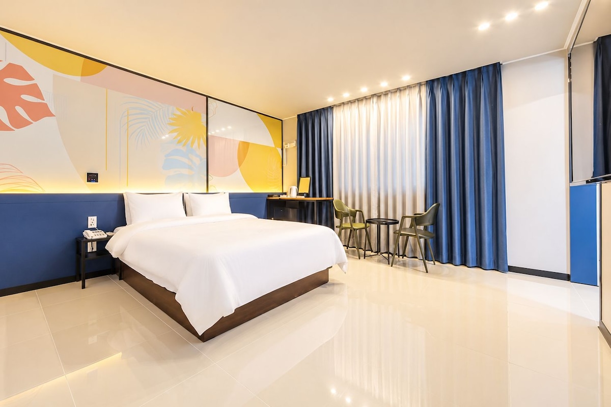 Quarto de hotel moderno com uma cama grande, uma escrivaninha com cadeira, uma mesa pequena com cadeiras e uma janela ampla com cortinas.