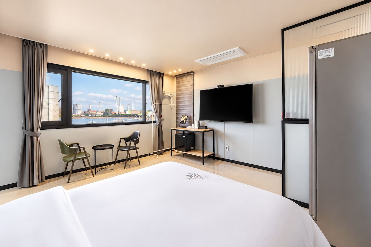 Quarto de hotel com cama, escrivaninha, cadeira, televisão e janela com vista para edifícios.