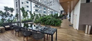 Uma área de estar moderna ao ar livre, com mesa de vidro e cadeiras de vime, cercada por vegetação exuberante e vista para um arranha-céu.