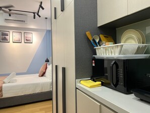 Quarto de hotel compacto com cama, micro-ondas e pequena cozinha equipada com pia e utensílios.