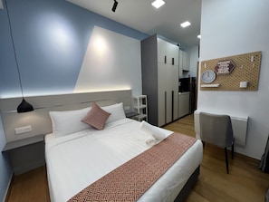Quarto de hotel moderno com cama grande, escrivaninha, cadeira, cozinha compacta e painel de rolhas na parede com frases motivacionais.