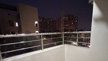 Uma varanda com guarda-corrimão de metal, chão branco e vista de edifícios altos à noite.