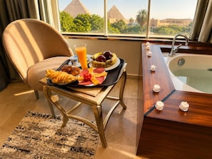 Bandeja de café da manhã com croissants, frutas e suco sobre uma mesa perto de uma janela com vista para pirâmides.