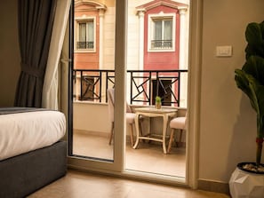 Quarto com cama, porta de vidro deslizante para uma varanda com mesa e cadeiras, e um vaso de planta.