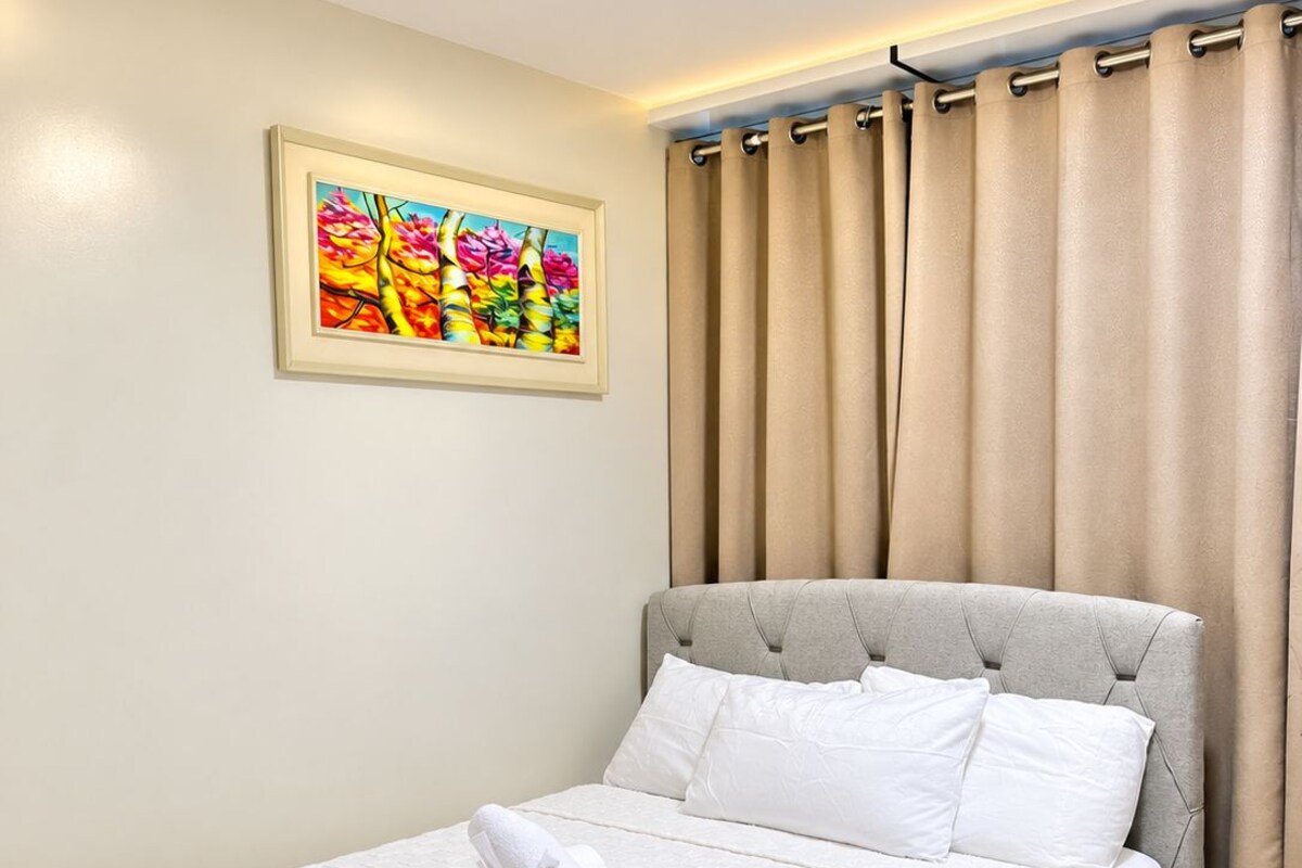 Cama bem arrumada com roupa de cama e almofadas brancas, um quadro na parede e uma janela com cortinas bege.