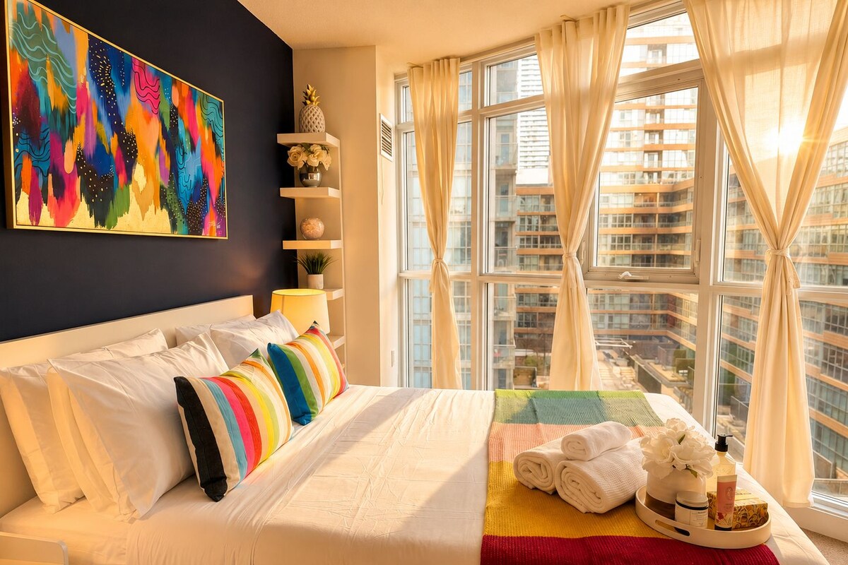 Quarto de hotel com cama grande, uma pintura colorida, vista para a cidade e bandeja com toalhas e artigos de higiene pessoal.
