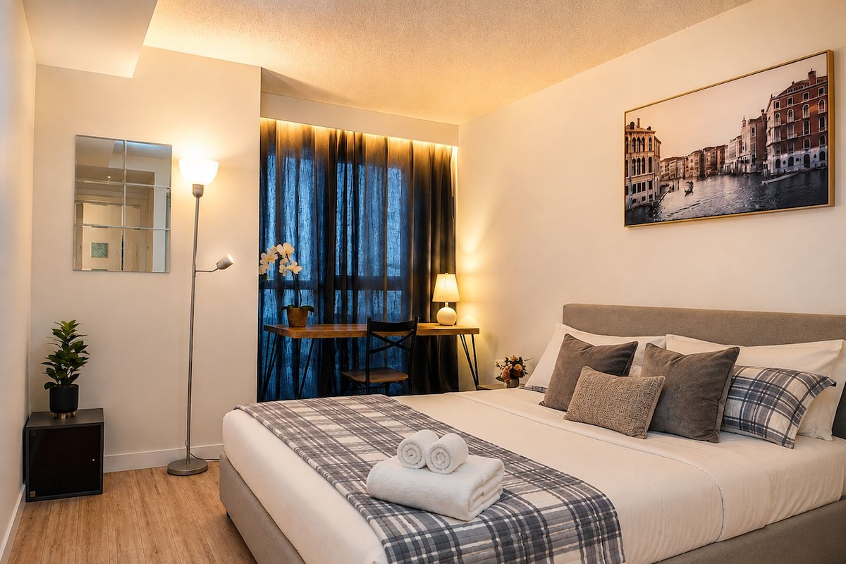 Quarto de hotel com cama, mesas de cabeceira, escrivaninha, cadeira, planta em vaso, abajur e um quadro na parede.