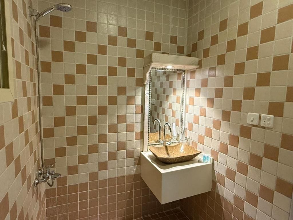 Casa de banho com padrão de azulejos xadrez, pia branca com cuba de madeira e chuveiro com chuveiro manual.