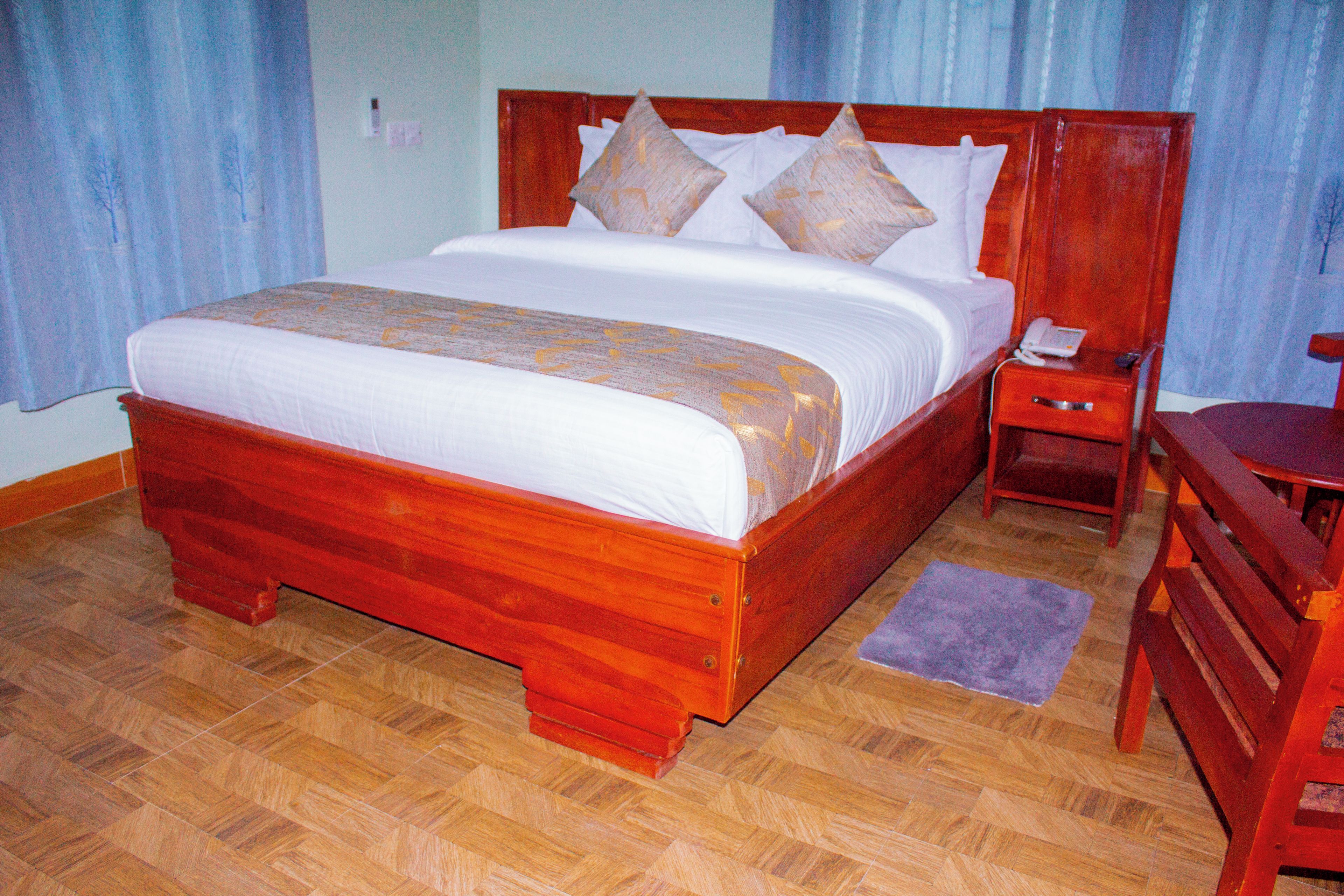 Uma cama de madeira com colchão branco e colcha estampada, uma mesa de cabeceira com telefone e um criado-mudo de madeira.