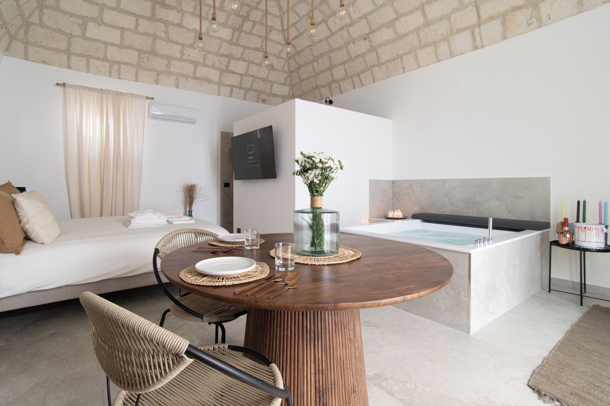 Quarto moderno com cama, mesa de jantar redonda, um vaso de vidro com flores, televisão de tela plana e uma banheira.