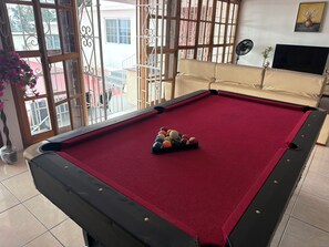 Uma mesa de bilhar com bolas, um sofá, uma televisão e uma janela com vista para edifícios.