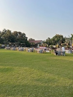 Um espaço ao ar livre para eventos, com várias mesas redondas e cadeiras, cercado por gramado e árvores.