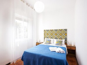 Quarto de hotel com cama, mesa de cabeceira, janela com cortinas leves e cabeceira estampada.