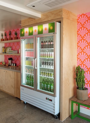 Geladeira para bebidas com várias prateleiras cheias de garrafas, um balcão de madeira com uma máquina de café e uma planta em vaso sobre uma mesa.