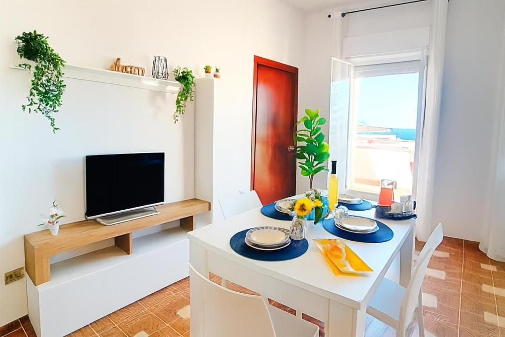 Uma área de refeições com uma TV em uma estante, uma mesa posta com pratos e um vaso de flores, e uma janela com vista.