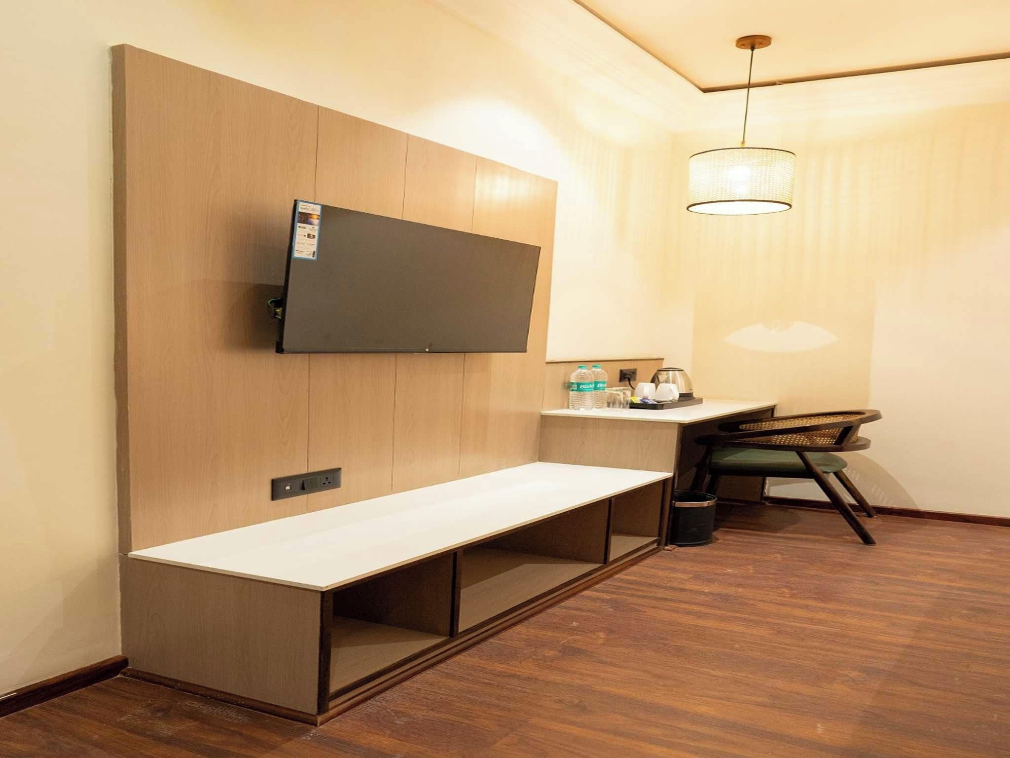 Quarto compacto com televisão integrada, um banco, uma mesinha com chaleira e uma cadeira.
