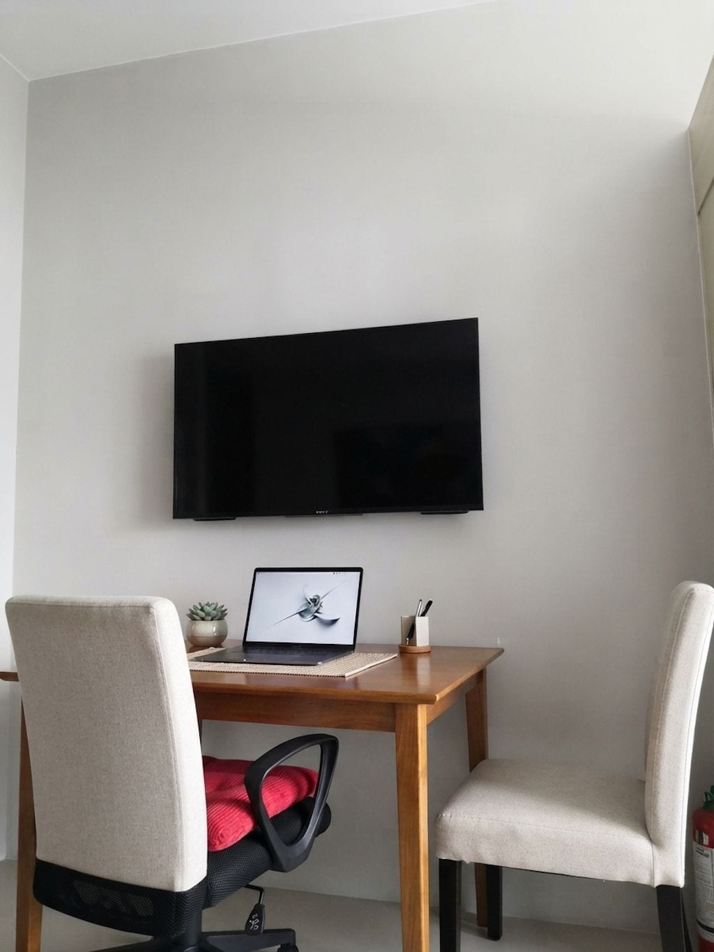 Quarto com televisão de tela plana na parede, escrivaninha de madeira com um laptop, duas cadeiras e uma pequena planta em vaso.