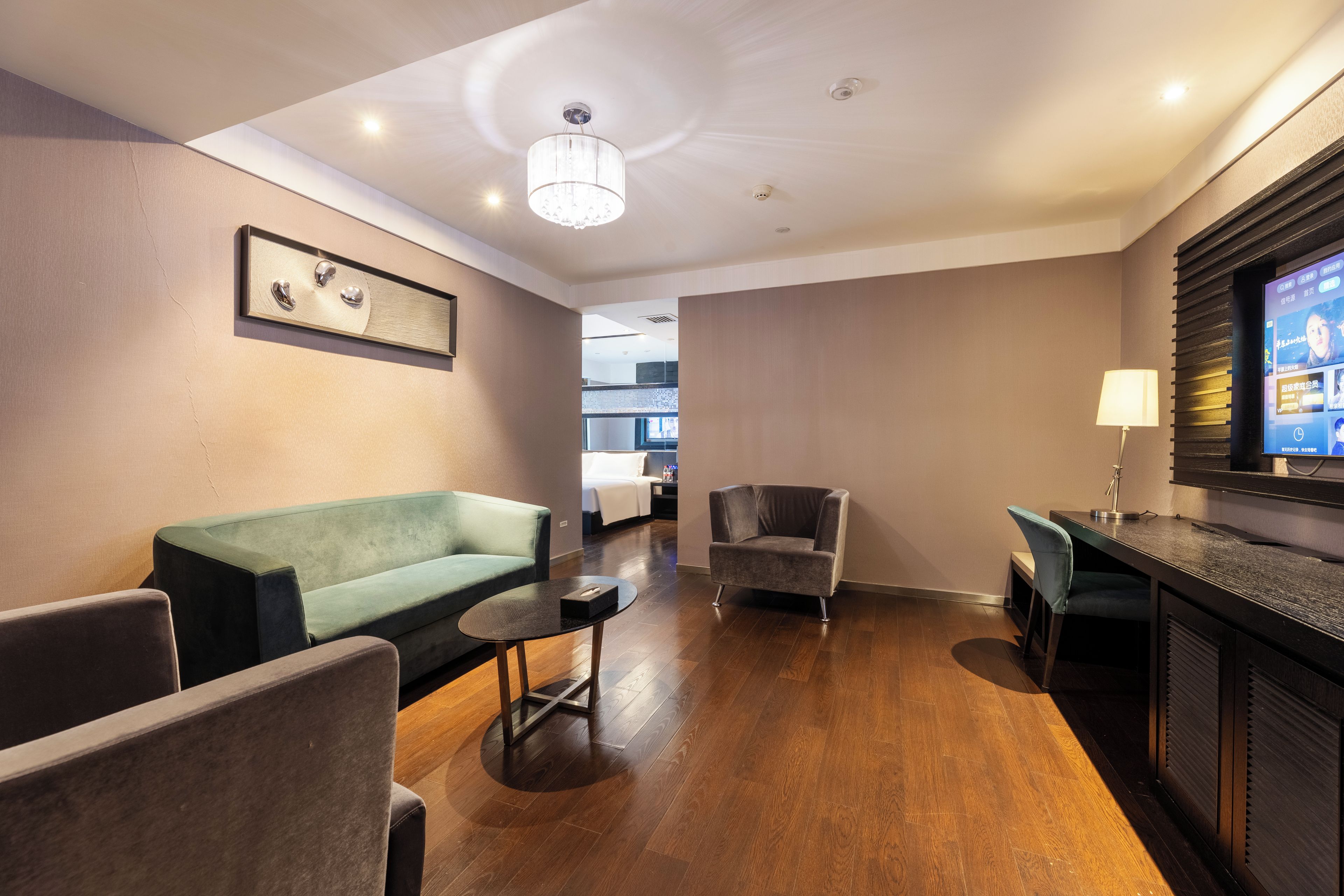 Quarto de hotel com sofá verde, uma mesa pequena, uma cadeira, televisão e piso de madeira.