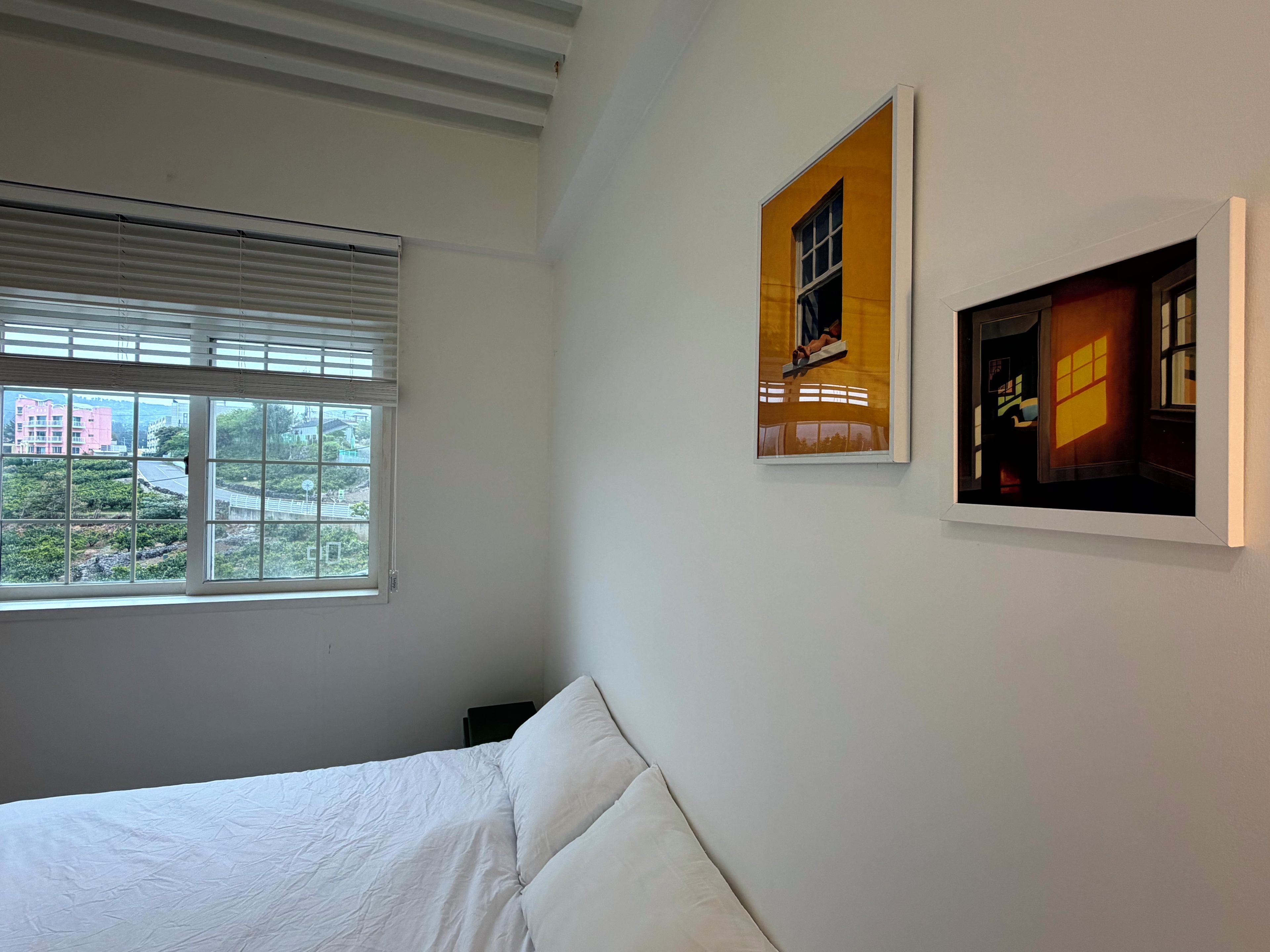 Quarto com cama, janela com persianas e duas fotos emolduradas na parede.