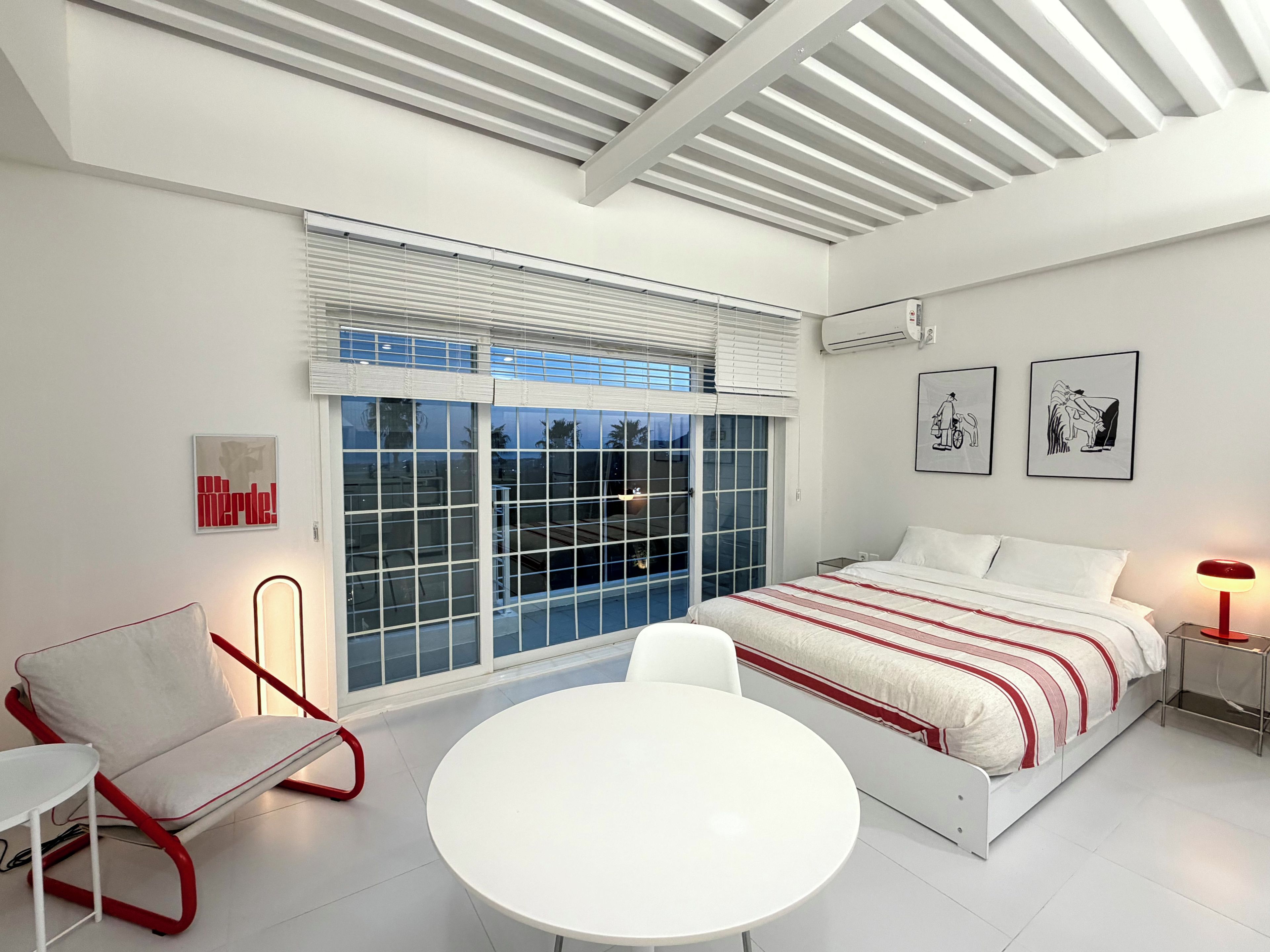 Quarto moderno com cama, mesa redonda, uma cadeira e uma janela grande com persianas.