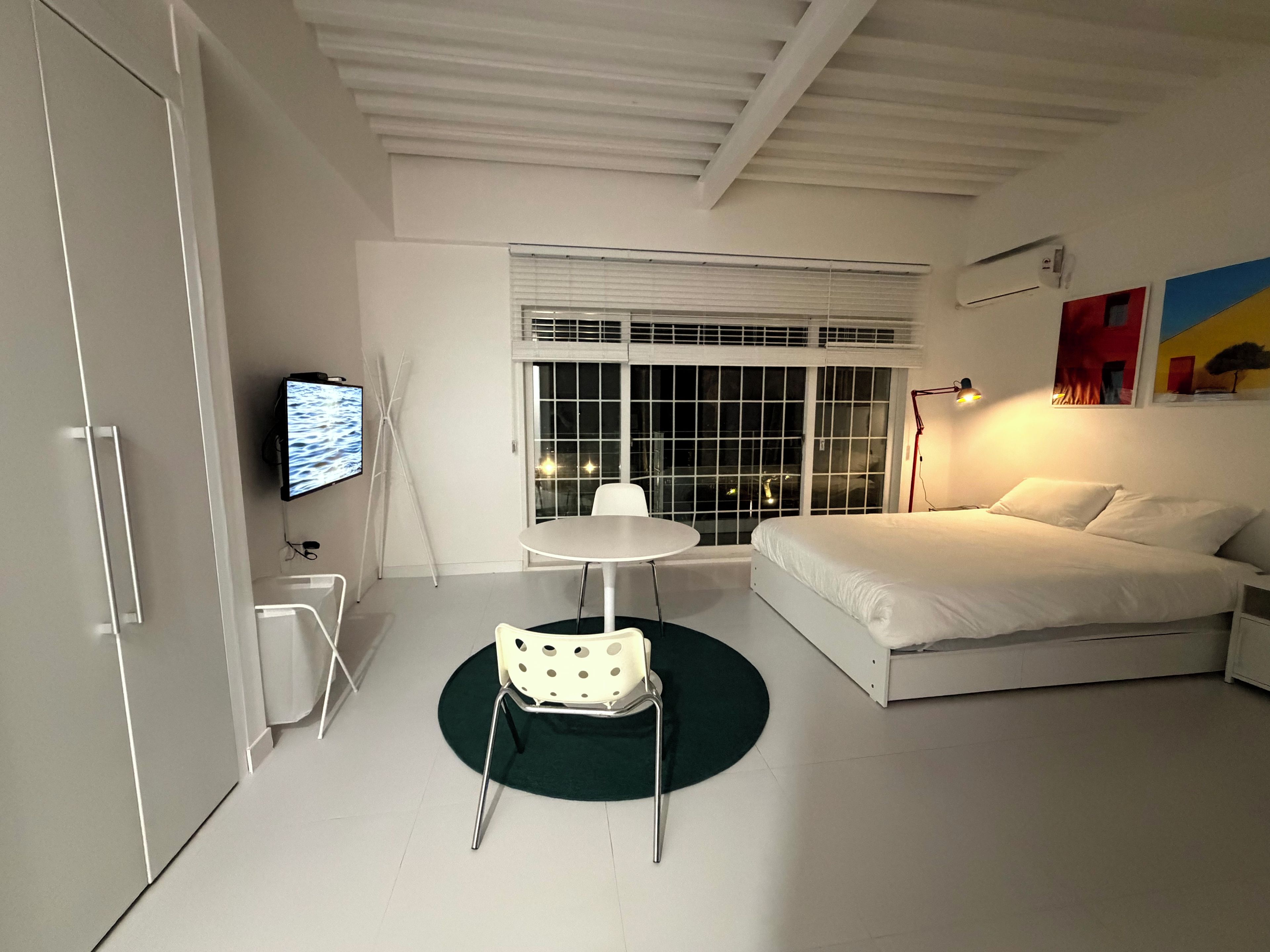 Quarto de hotel moderno com uma cama, uma mesinha, uma cadeira, uma televisão e uma janela com persianas.