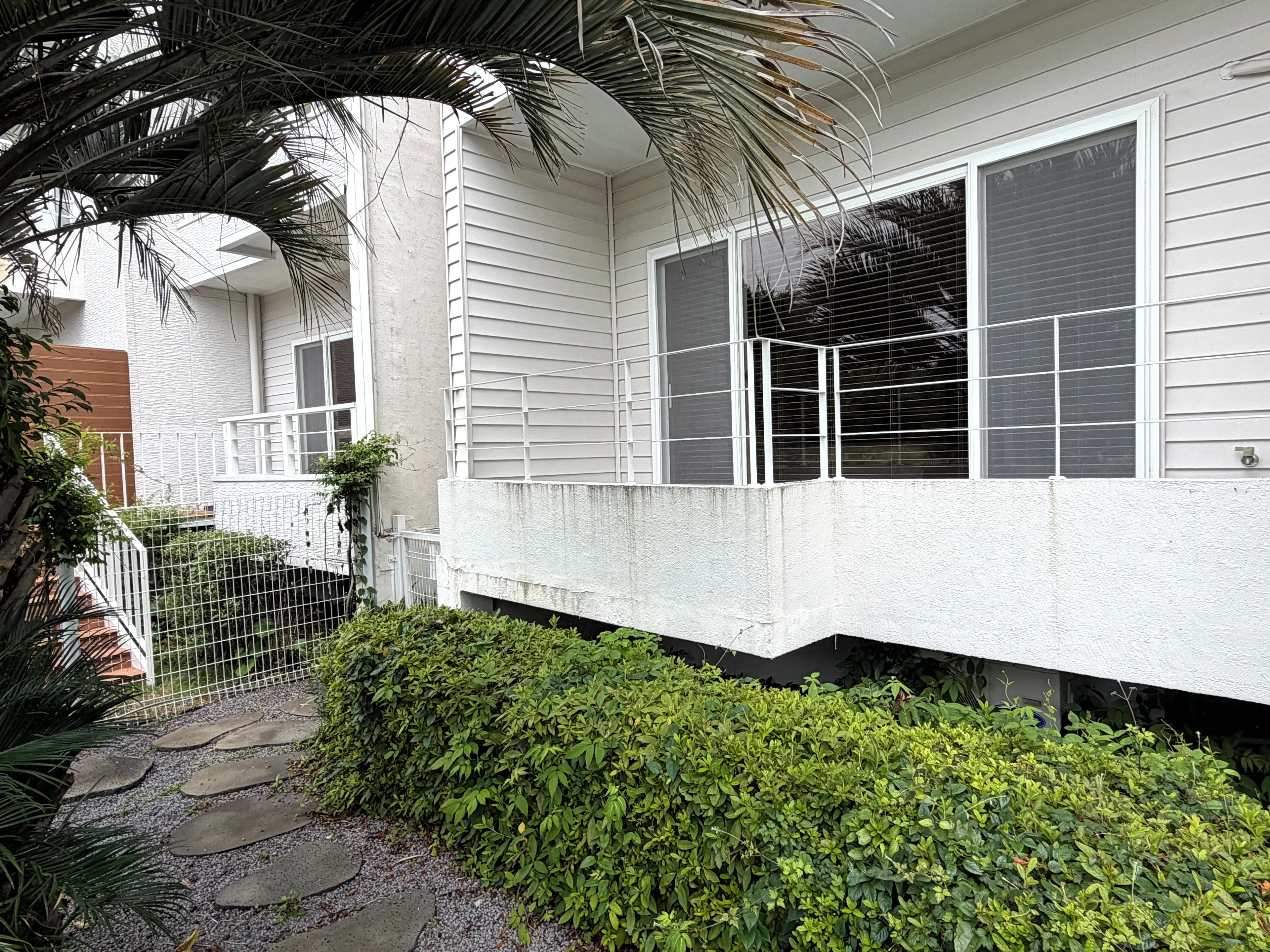 Edifício residencial com fachada branca, entrada protegida por portão e cerca viva bem cuidada.