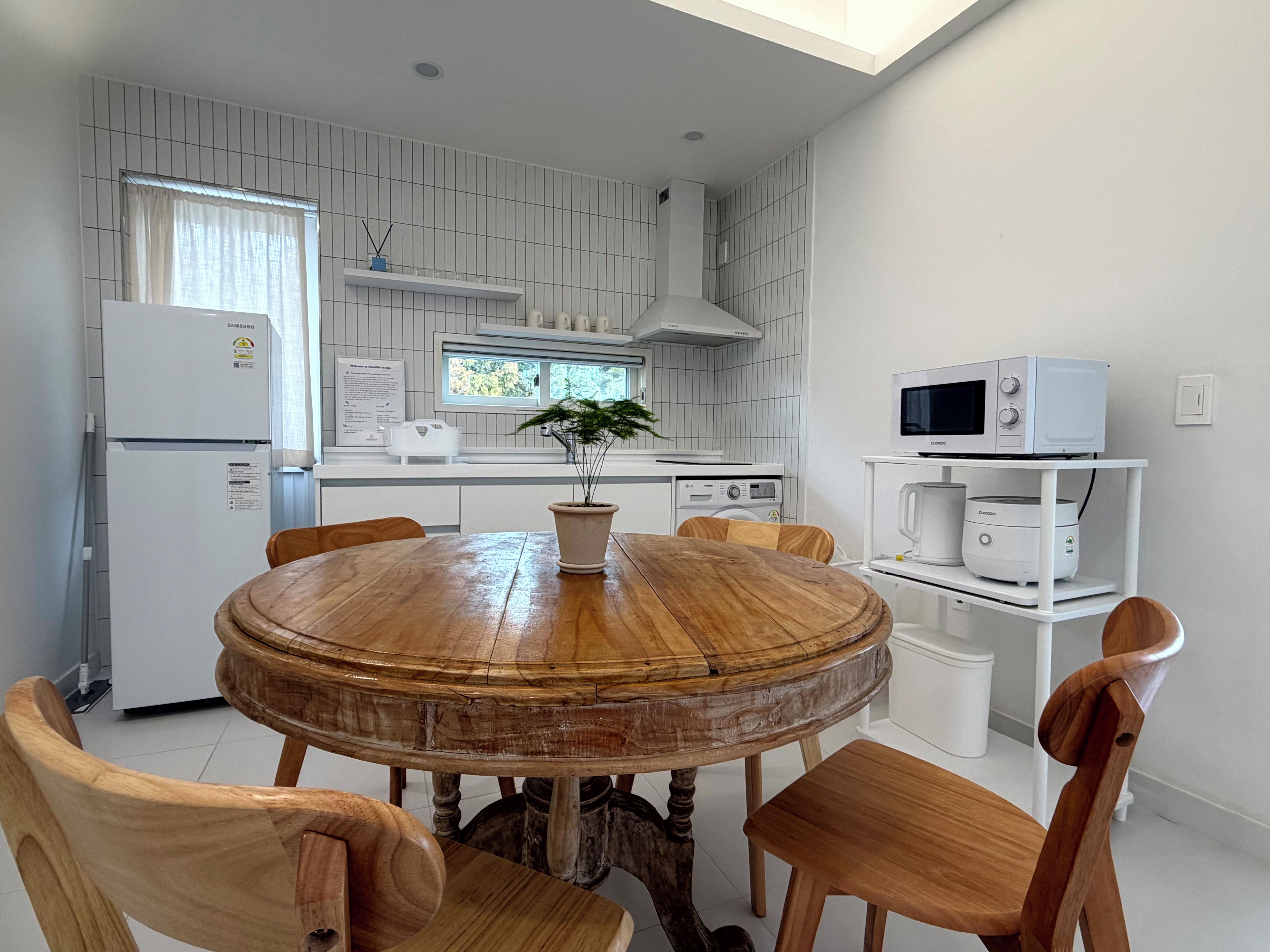 Uma cozinha moderna com uma mesa de madeira redonda, armários brancos, micro-ondas embutido e uma geladeira Samsung.