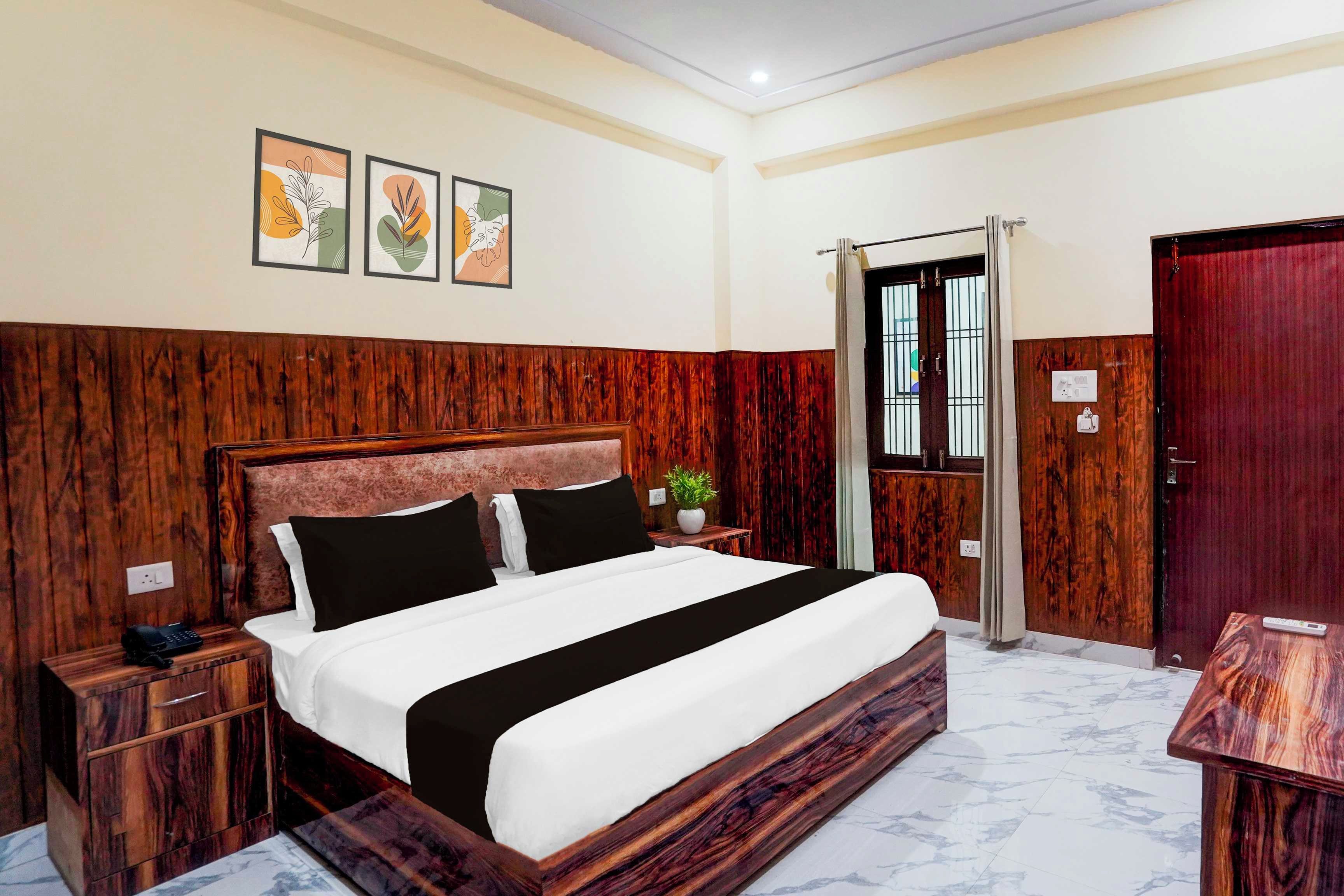 Quarto de hotel com cabeceira de madeira, cama com roupa de cama branca, mesa de cabeceira e mesa lateral com uma planta, e porta com janela.